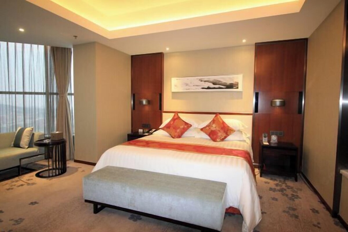 Quarto de hotel com uma cama grande, um banco, um sofá, uma mesinha e um quadro na parede.