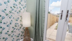 Um quarto com papel de parede estampado, um candeeiro de chão trançado, uma cortina verde e uma porta que dá para o exterior.