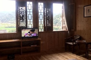 Quarto com paredes de madeira e uma TV sobre um móvel de madeira. Há janelas com telas decorativas, uma cadeira e uma pequena mesa.