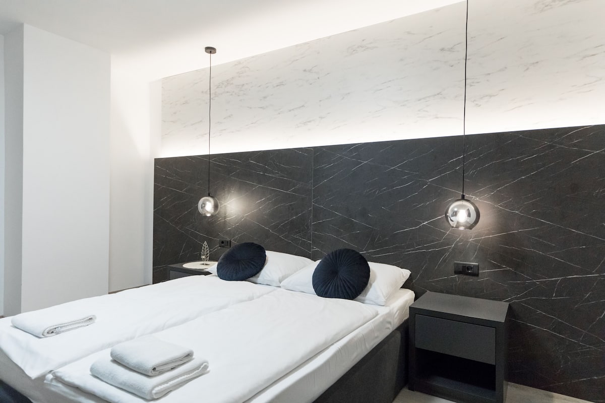 Quarto moderno com cama grande, cabeceira de mármore escuro, mesas de cabeceira e iluminação embutida na parede.