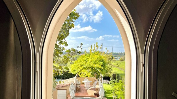 Visão através de uma janela com arco gótico, mostrando uma área de piscina com estátuas, um jardim e céu claro.