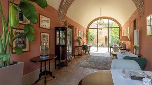 Uma área de refeições com teto alto abobadado, uma janela grande e vista para o exterior. Há mesas postas para refeições, um candelabro e um vaso de planta.