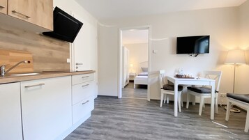 Cozinha moderna com televisão integrada, sala de jantar com um tabuleiro de xadrez e um quarto visível através de uma porta aberta.
