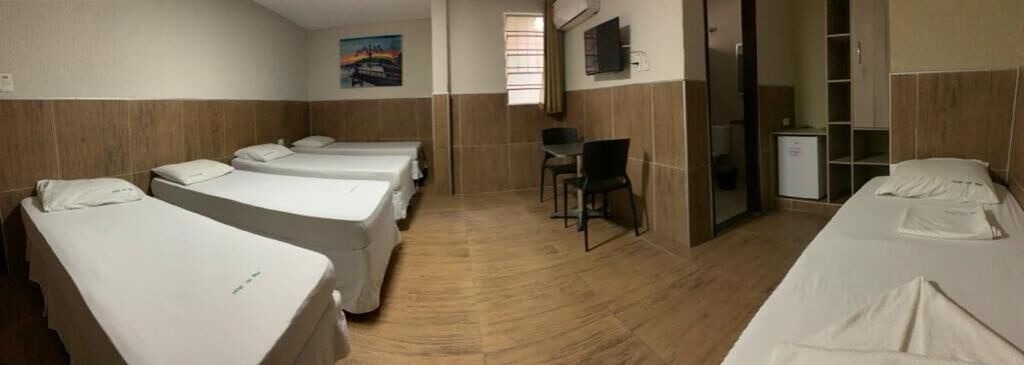Quarto de hotel com quatro camas de solteiro, uma mesinha, cadeiras e uma televisão fixada na parede.