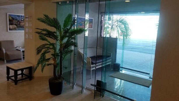 Uma área de recepção com divisória de vidro, um vaso de planta, uma cadeira de vime e uma pequena mesa.