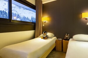 Quarto de hotel com duas camas de solteiro, uma janela com vista para uma paisagem nevada, um abajur na parede e um pinguim de pelúcia em uma das camas.