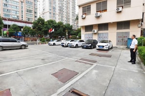 Um estacionamento com vários carros, um edifício com aparelhos de ar condicionado e uma pessoa parada perto da entrada.
