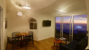 Uma sala de estar com piso de madeira, uma mesa de jantar redonda com cadeiras, um sofá, uma TV fixada na parede e uma grande porta de vidro deslizante com vista para o céu.