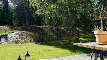 Um deck de madeira com mesa e cadeiras, cercado por um gramado com árvores e balanço.