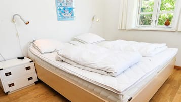 Cama bem arrumada com roupa de cama branca, mesa de cabeceira com abajur e janela com vista para o verde.