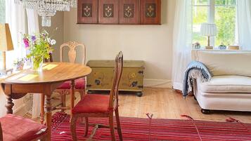 Um quarto com uma mesa de jantar, cadeiras, um sofá, um tapete estampado, um candelabro e uma janela com cortinas.