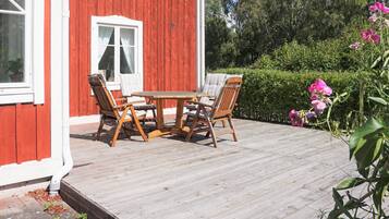 Um deck de madeira com mesa e cadeiras, um edifício vermelho com detalhes brancos, e um jardim com flores cor-de-rosa.