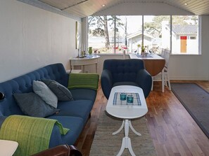 Quarto com sofá azul, mesa branca com dois copos e vista para uma casa através de janelas amplas.