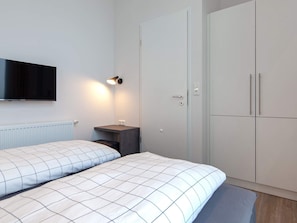 Quarto com duas camas, televisão na parede, mesa de cabeceira e guarda-roupa.
