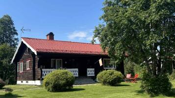 Casa de madeira com telhado vermelho, varanda e uma bandeira. Arbustos e gramado à frente.