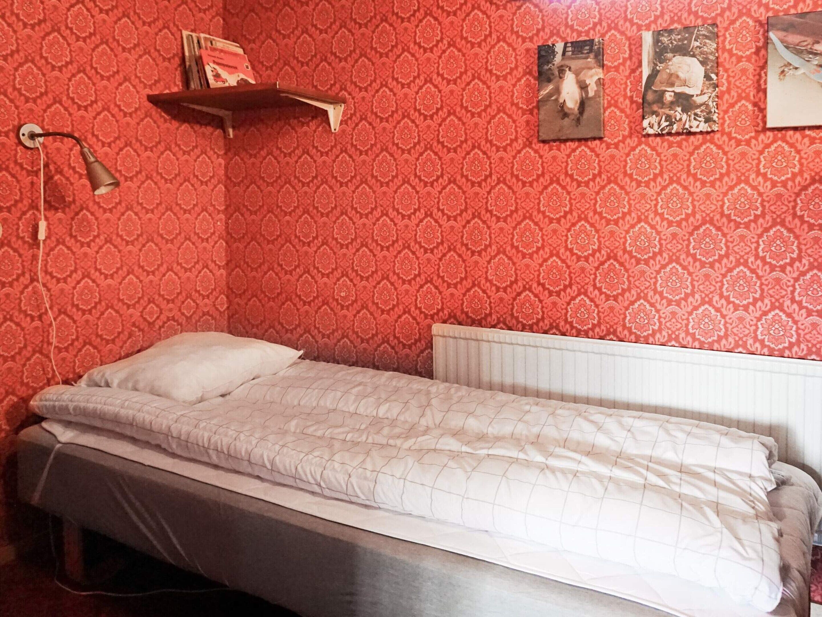 Uma cama de solteiro com colcha branca num quarto com papel de parede estampado em vermelho.