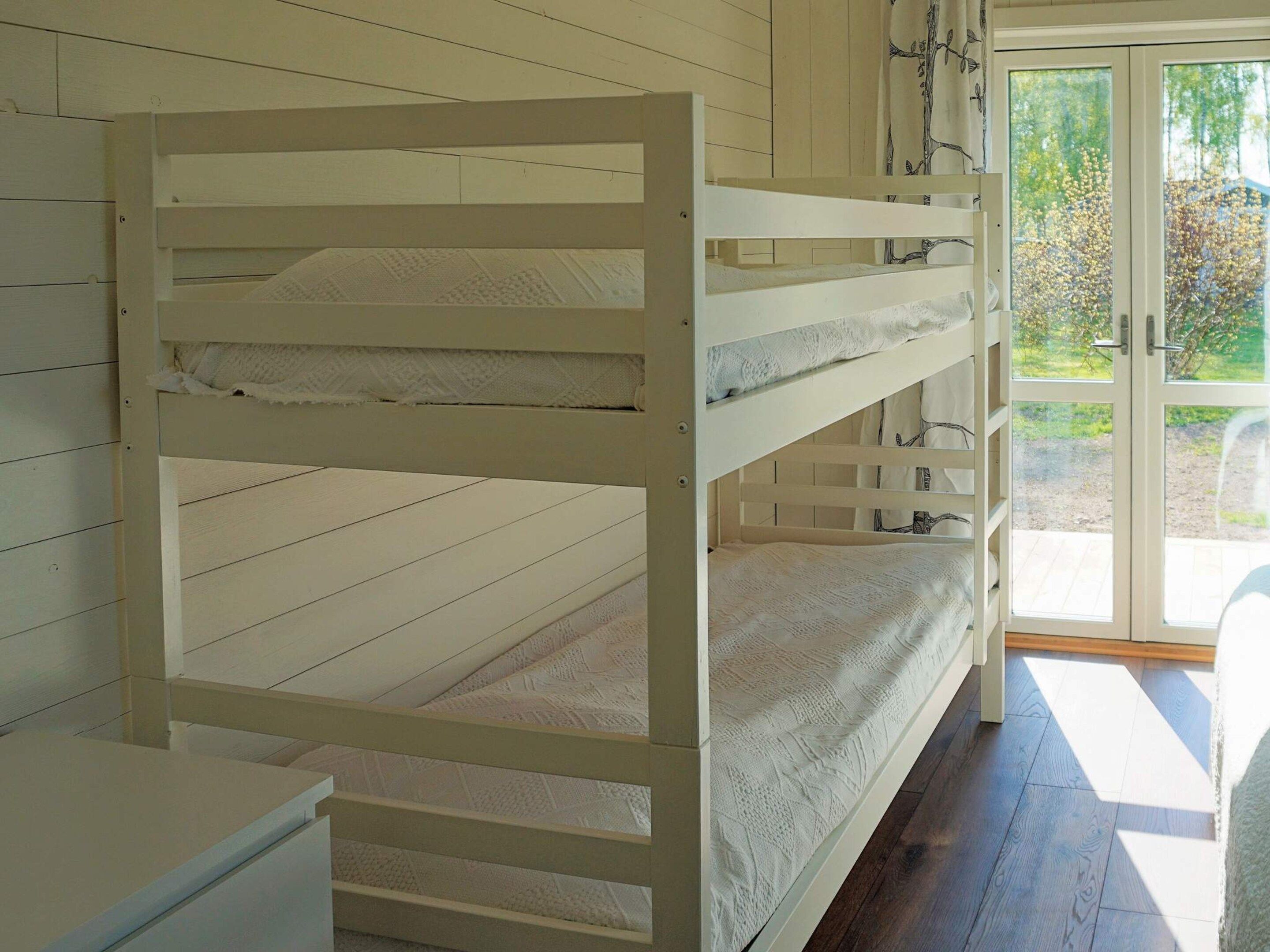 Uma cama de solteiro com estrutura de madeira e roupa de cama branca, uma cômoda pequena e branca, e uma porta de vidro corrediça que dá para uma área externa.