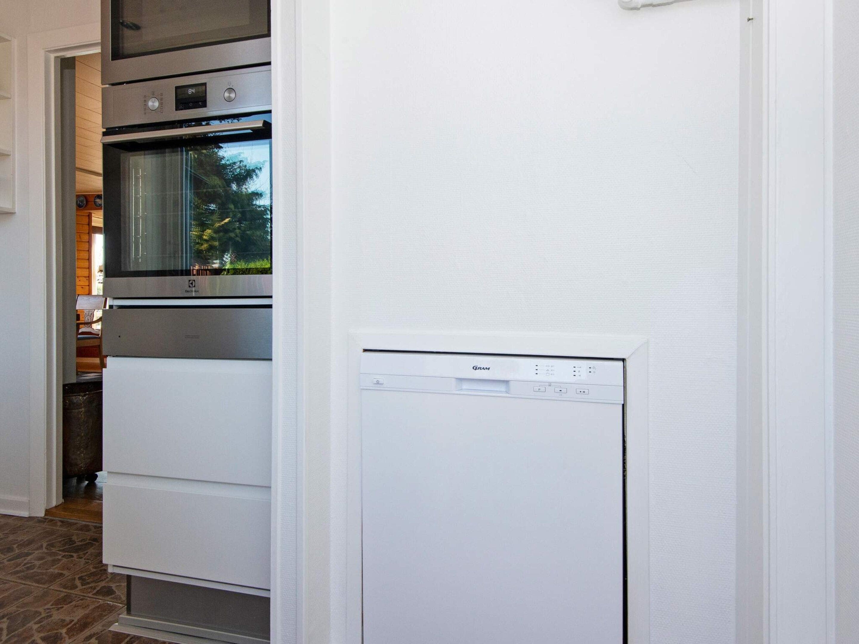 Cozinha com forno embutido e máquina de lavar louça, janela com vista para o verde e porta que dá para outro cômodo.