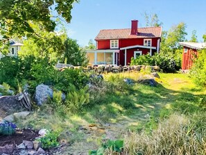 Uma casa vermelha com telhado inclinado, cercada por um jardim com diversas plantas e um muro de pedra.