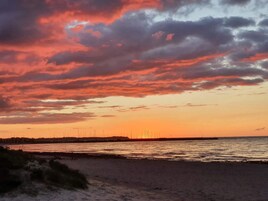 Uma praia com um pôr do sol, um píer ao longe e céu claro.
