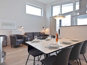 Uma área de refeições moderna com uma mesa branca posta para quatro pessoas, um sofá cinza e uma janela grande que proporciona luz natural.