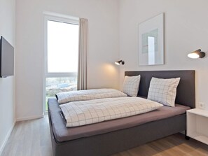 Quarto moderno com cama, mesa de cabeceira, luminária na parede e janela com cortinas.