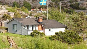 Uma pequena casa com chaminé, uma bandeira e um morro rochoso ao fundo.
