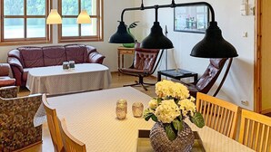 Uma área de jantar com uma mesa posta para quatro pessoas, rodeada por cadeiras de madeira. Há um vaso com flores sobre a mesa, e uma pequena mesa com uma lâmpada e uma cadeira ao fundo.