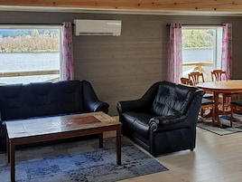 Quarto aconchegante com sofá, mesa de centro e mesa de jantar com cadeiras. Há uma janela com cortinas, um aparelho de ar condicionado na parede e vista para um corpo d'água lá fora.