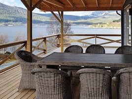 Um terraço coberto ao ar livre com uma mesa e cadeiras de vime, com vista para um lago e montanhas.