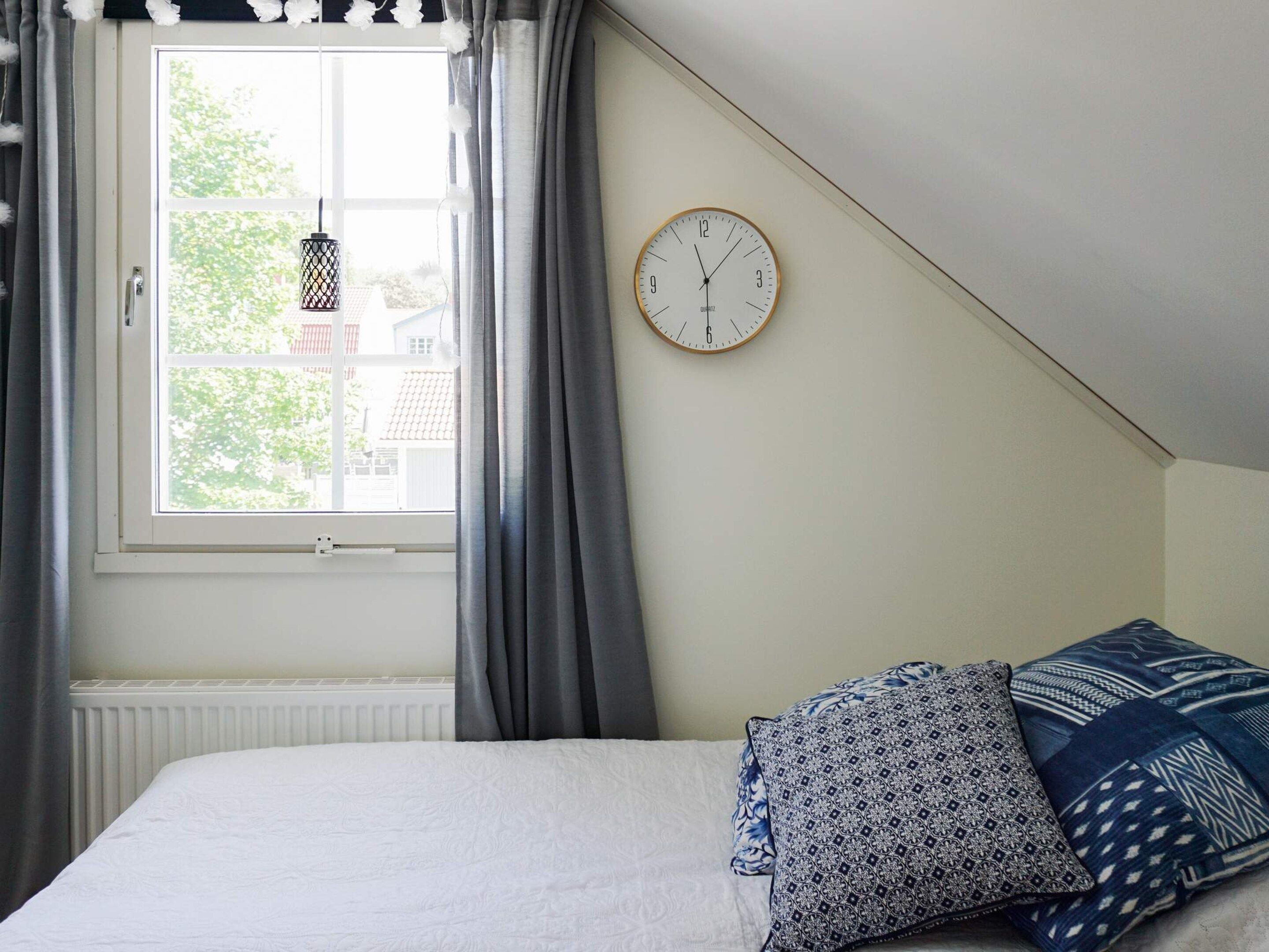 Um quarto com cama, janela com cortinas, relógio na parede e radiador.