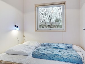 Quarto com uma cama de solteiro, janela com vista para árvores secas e luminária na parede.