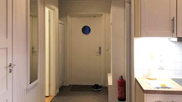 Um corredor estreito que leva a uma porta com abertura circular, um extintor fixado na parede e um pequeno tapete no chão.