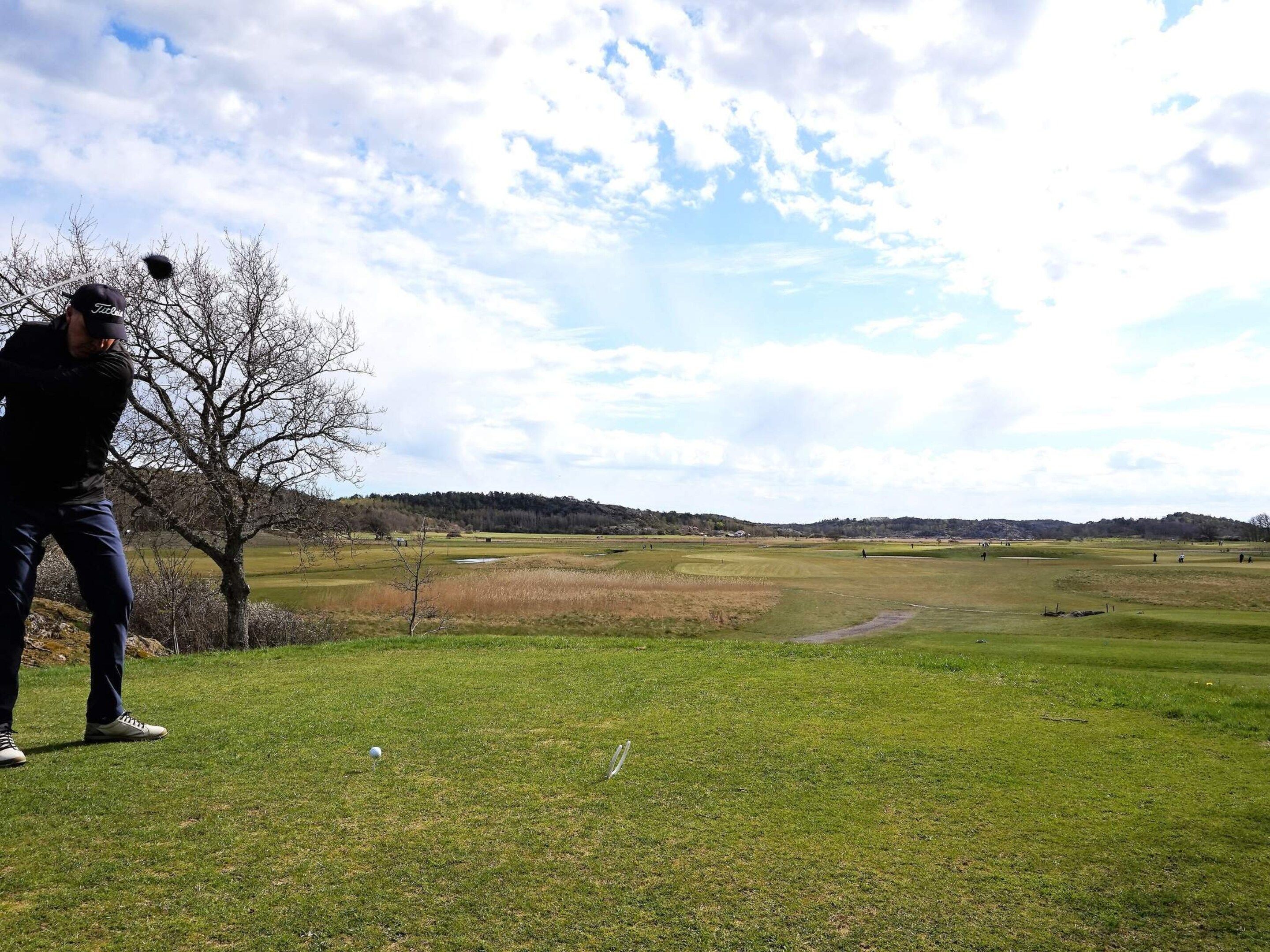 Campo de golfe com uma pessoa se preparando para golpear a bola, árvores e céu claro.