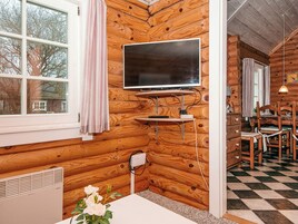 Interior acolhedor de uma cabana, com uma televisão de tela plana montada, paredes de madeira, área de jantar com piso xadrez e um radiador.