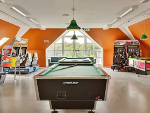 Mesa de bilhar numa sala com paredes laranja, jogos de fliperama e vista através de grandes janelas.