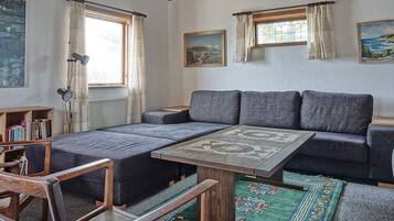 Quarto aconchegante com sofá, mesa de madeira e uma cadeira. Há estantes de livros e um abajur.