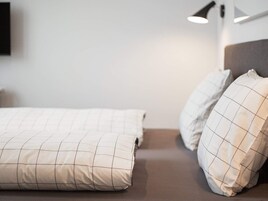 Uma cama bem arrumada com colcha e almofadas quadriculas em branco e bege, cabeceira cinza e luminária fixada na parede.