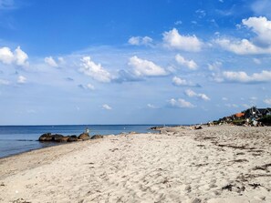 Uma praia arenosa com pedras espalhadas, céu azul límpido e alguns edifícios ao fundo.