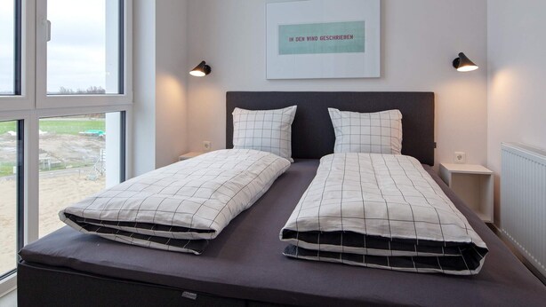 Uma cama de casal com cabeceira, duas mesas de cabeceira com luminárias, uma frase emoldurada na parede e vista para o exterior por uma grande janela.