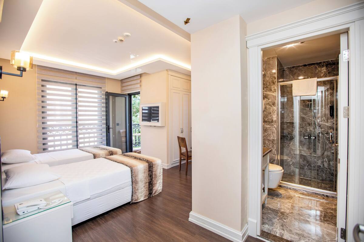 Quarto moderno com uma cama grande, uma mesa de cabeceira e um armário de vidro. Dá para ver um banheiro com chuveiro e vaso sanitário através de uma porta de vidro.