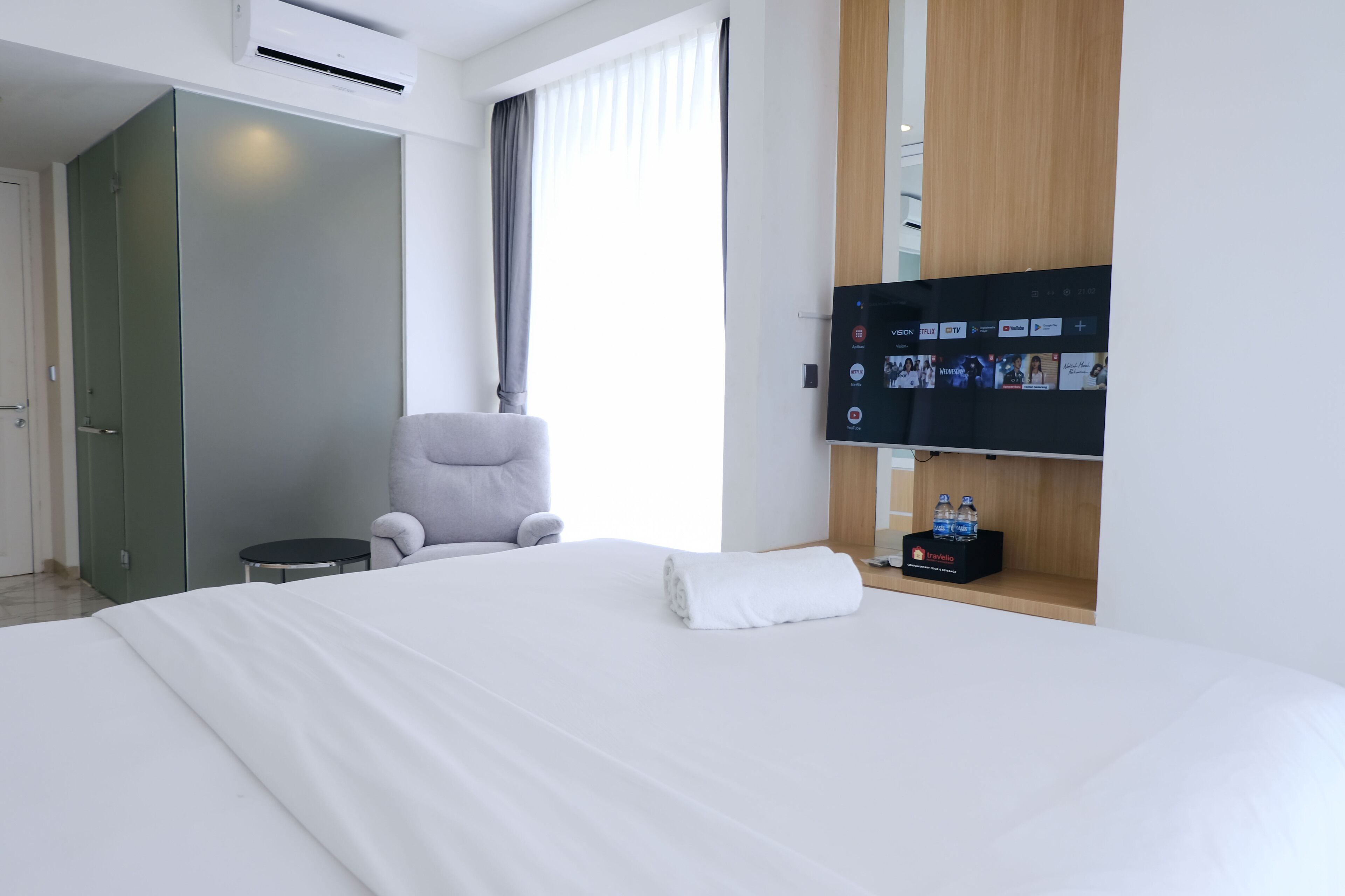 Quarto de hotel moderno com uma cama grande, uma poltrona cinzenta, uma televisão na parede e um minibar.