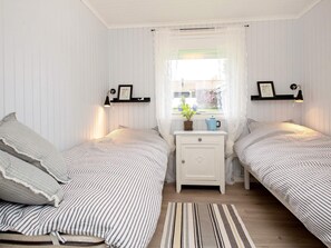 Quarto com duas camas de solteiro, mesa de cabeceira com abajur, janela com cortinas e uma pequena estante com uma planta e um quadro.