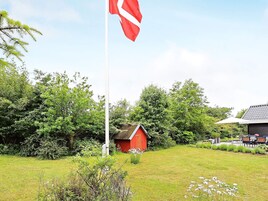 Um edifício vermelho com cerca branca, rodeado por vegetação e um mastro com uma bandeira.