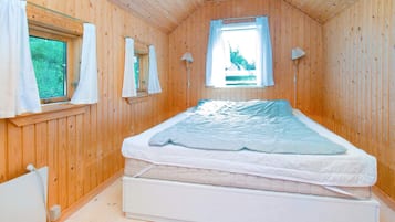 Um quarto pequeno, com painéis de madeira, uma cama, duas janelas com cortinas e teto inclinado.