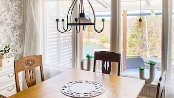 Uma sala de jantar com mesa de madeira, cadeiras, um candelabro e vista para o exterior através de grandes janelas.