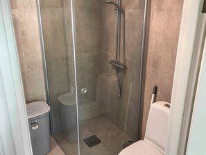 Divisória de chuveiro com paredes de vidro, chuveiro manual, vaso sanitário e cesto de lixo.