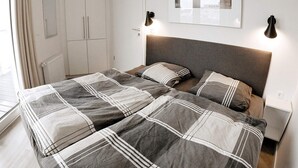 Uma cama de casal com colcha cinza e branca quadriculada, cabeceira cinza e duas luminárias de cabeceira.