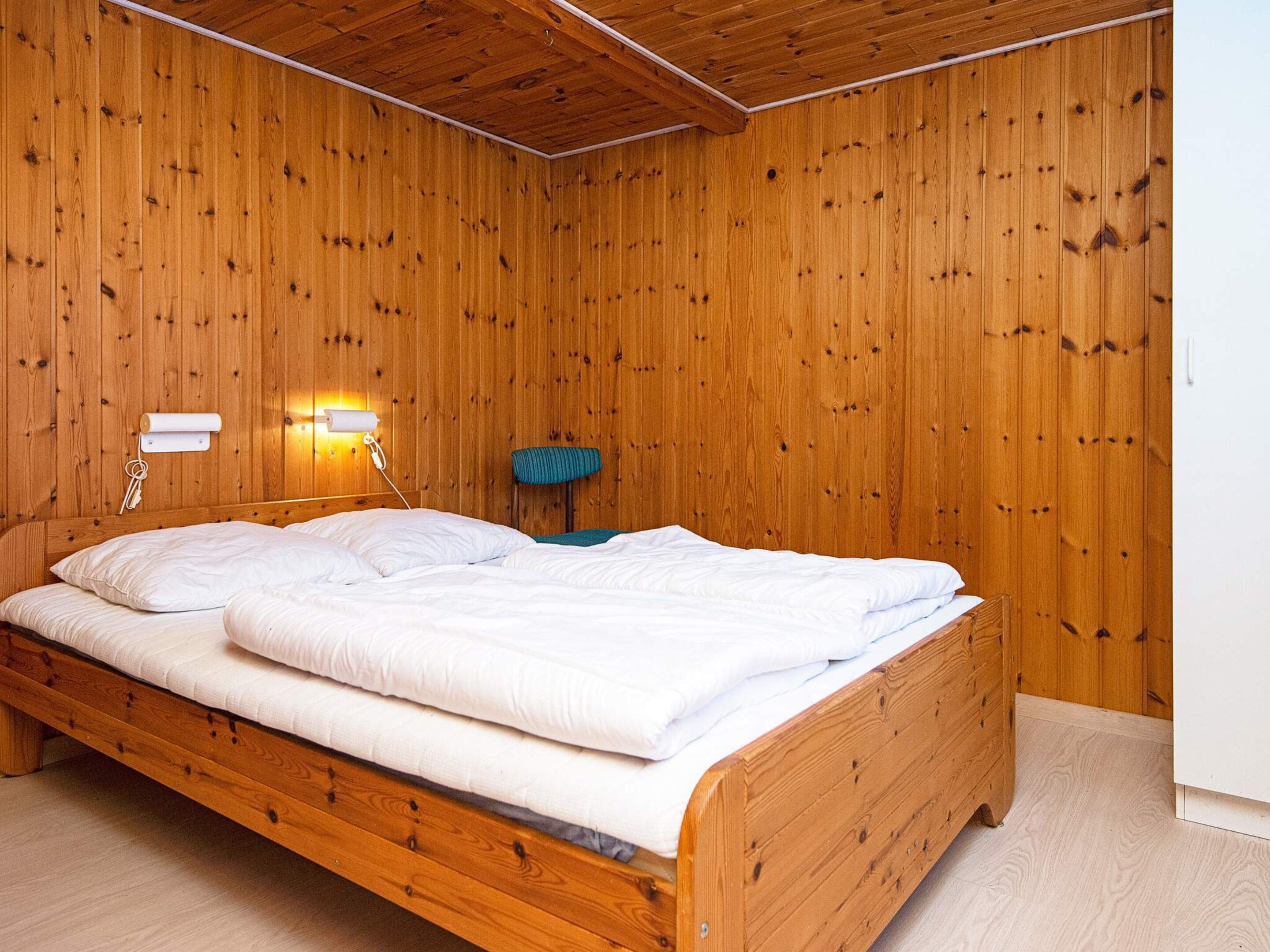 Quarto com painéis de madeira, uma cama, luminária na parede e uma cadeira.