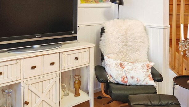 Uma sala de estar com uma TV de tela plana sobre um móvel branco, uma poltrona macia com almofada floral, um abajur preto e uma escada de madeira.